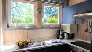 Cozinha com pia, fogão e micro-ondas. Janelas oferecem vista para árvores.