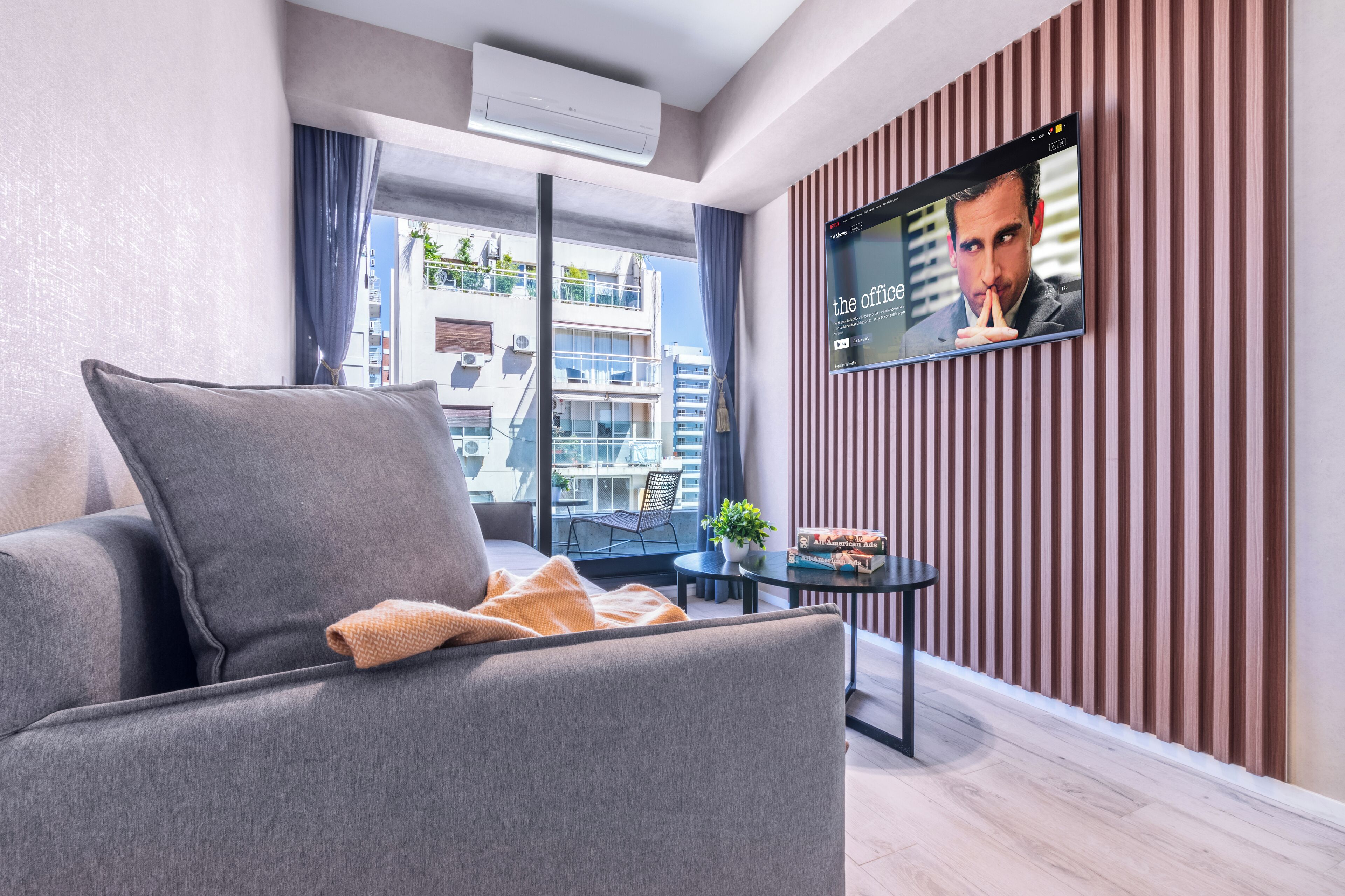 Uma sala de estar moderna com um sofá cinzento, uma pequena mesa redonda com uma planta e um jogo de tabuleiro, uma TV fixada numa parede riscada, e uma varanda com vista para edifícios.