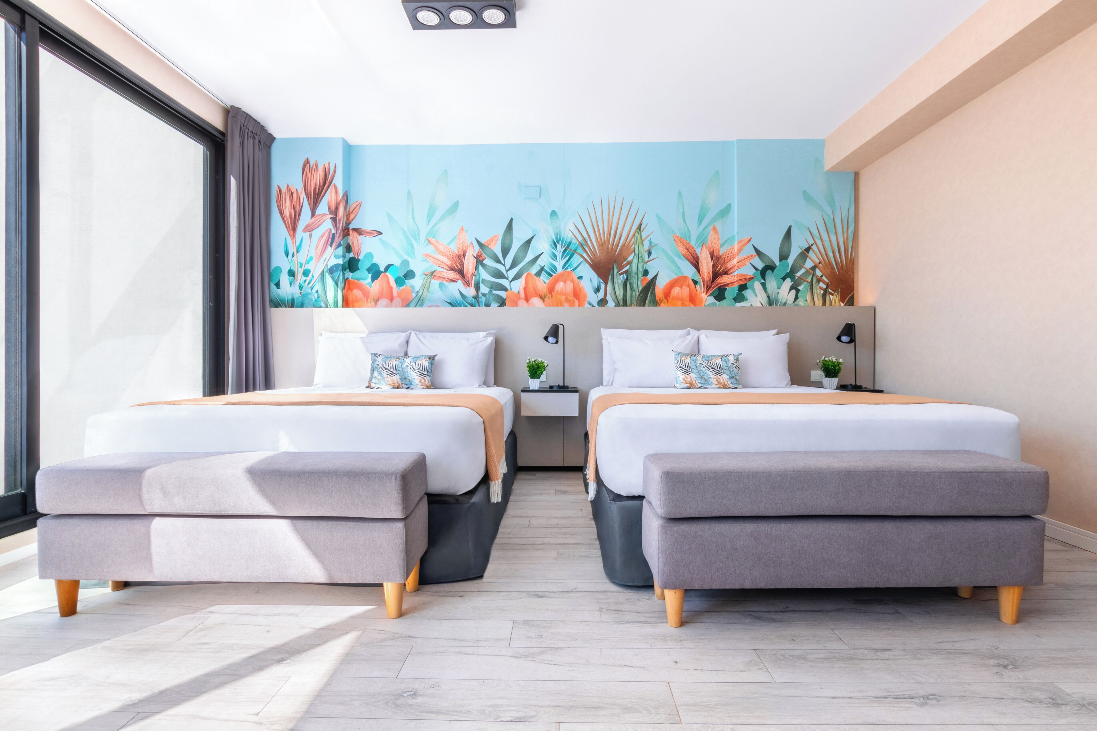 Quarto de hotel moderno com duas camas, uma janela grande e um mural de plantas tropicais na parede.