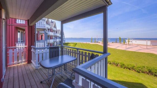 Um deck de madeira com mesa e cadeiras, com vista para uma praia e um edifício com fachadas vermelhas e brancas.