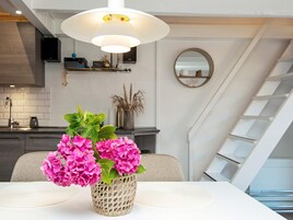 Uma cozinha moderna com uma luminária branca no teto, uma mesa de jantar e um cesto de flores rosa, e uma escada com corrimão branco.