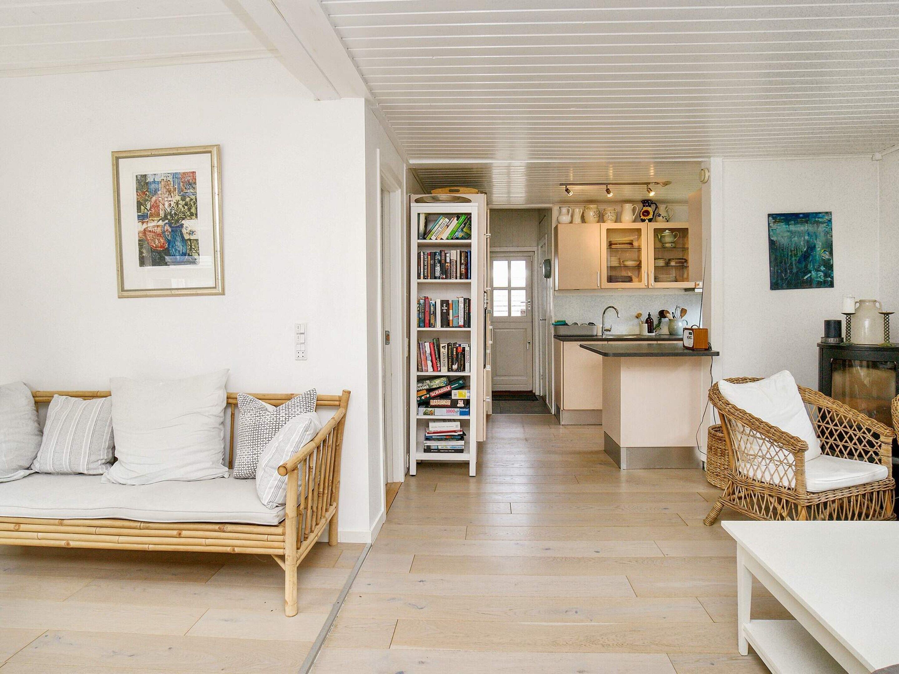 Uma sala de estar aconchegante com um sofá e uma poltrona de vime, uma estante repleta de livros e um espaço de cozinha com armários e pia.