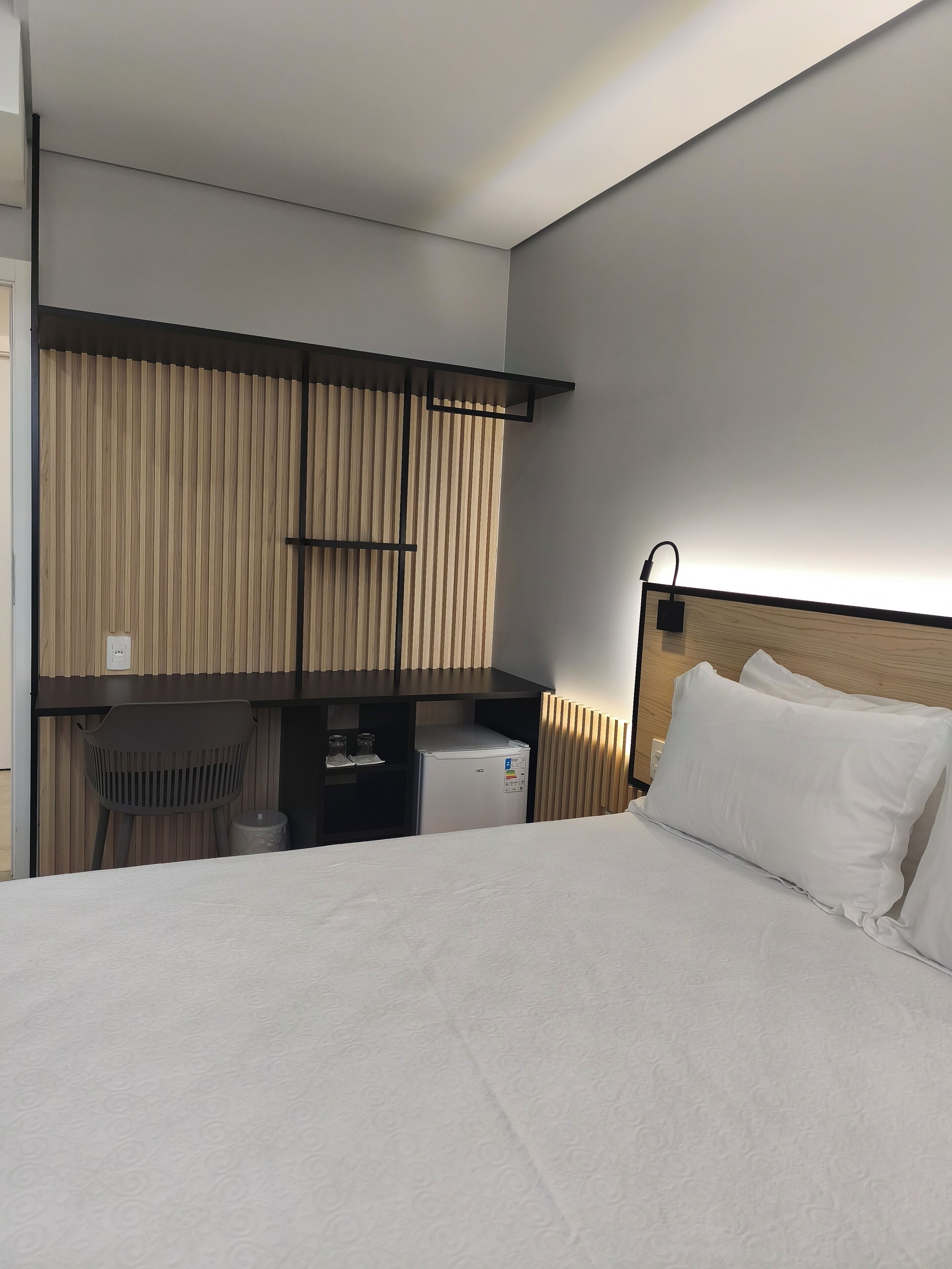 Quarto de hotel com cama, escrivaninha, cadeira e um pequeno frigorífico.