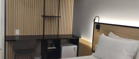 Quarto de hotel com cama, escrivaninha, cadeira e um pequeno frigorífico.