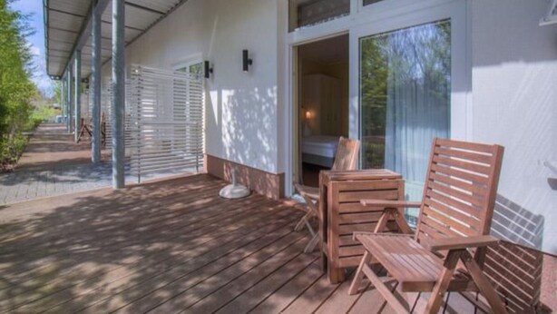 Um deck de madeira com móveis de exterior, uma área coberta e um quarto visível através de uma janela.