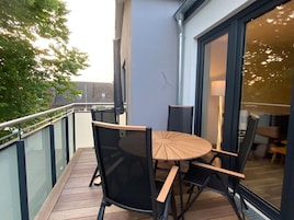 Uma varanda com mesa redonda e cadeiras, guarda-corpo de vidro e vista para a paisagem urbana.