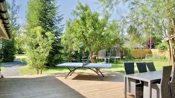 Um deck externo com mesa de pingue-pongue e cadeiras de vime, rodeado por árvores e um gramado.