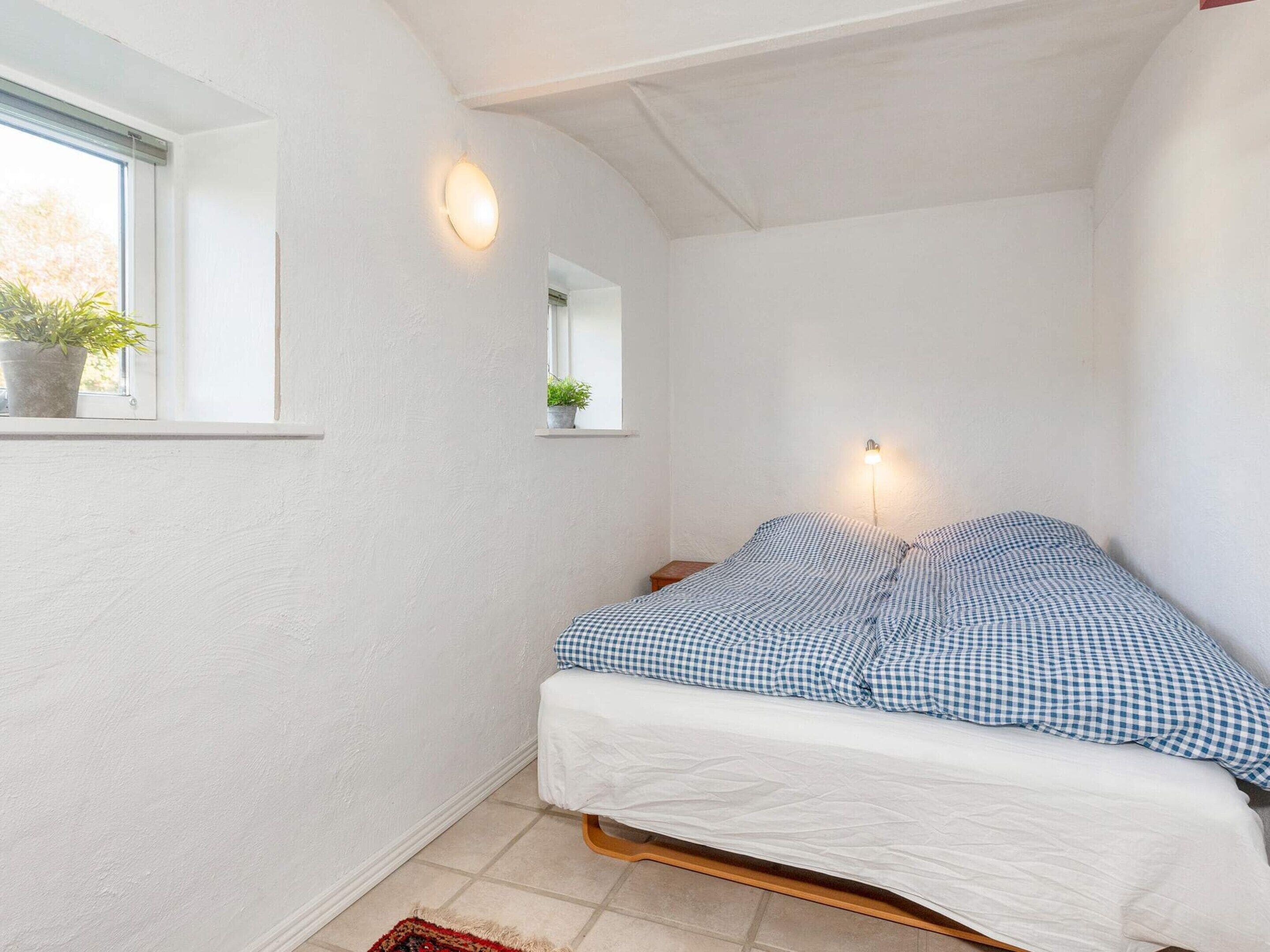Um quarto pequeno com uma cama de solteiro, uma janela com uma planta e uma luminária na parede.