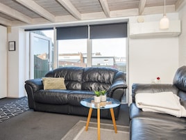 Uma sala de estar moderna com um sofá de couro preto, uma mesa de centro de vidro com duas plantas, e vista para o exterior através de uma porta de correr de vidro.