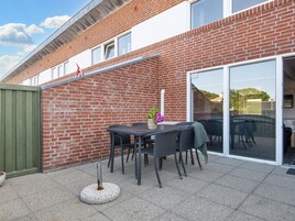 Uma área de pátio com mesa e cadeiras, uma pequena mesa redonda com uma planta e uma porta de vidro deslizante que dá para um espaço ao ar livre coberto.
