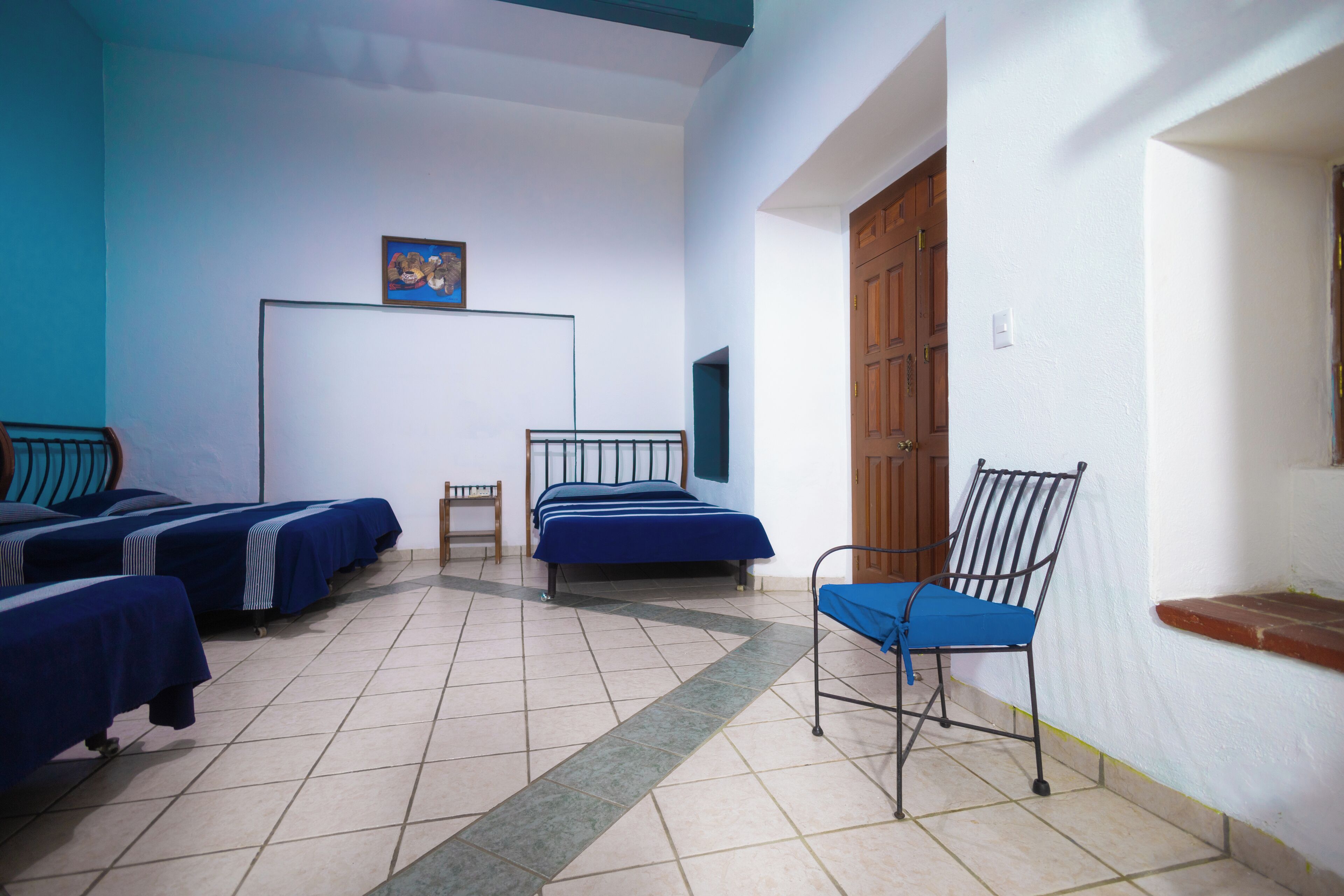Quarto com três camas, uma cadeira, porta de madeira e um quadro na parede.