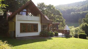 Uma casa de dois andares com varanda, cercada por vegetação exuberante e um lago.