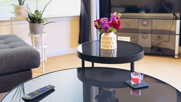 Uma sala de estar moderna com uma mesa de centro de vidro, um controle remoto, um vaso com flores e uma TV ao fundo.