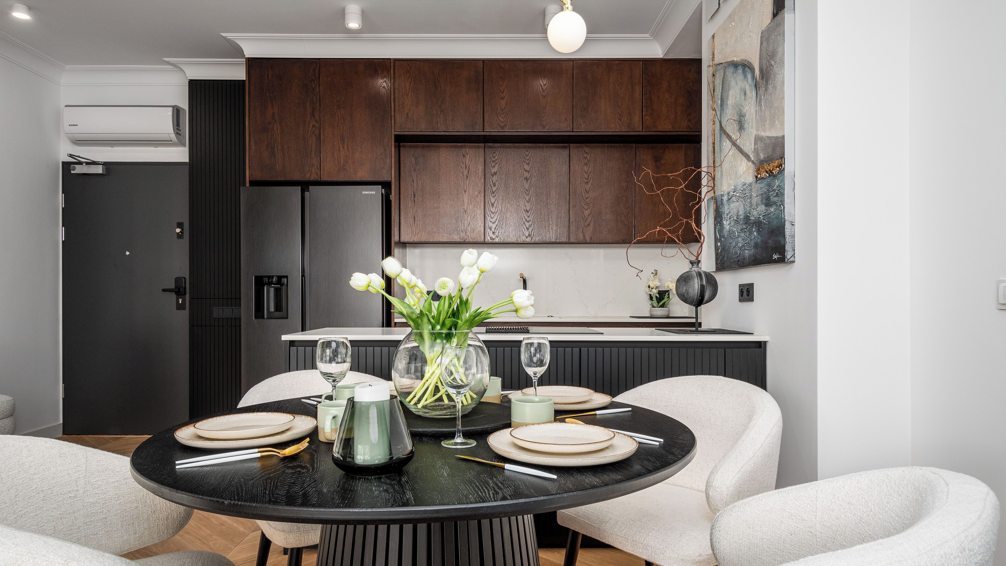 Uma área de refeições moderna com uma mesa redonda posta para quatro pessoas, rodeada por cadeiras brancas. Há um vaso com flores brancas sobre a mesa, e uma cozinha visível ao fundo, com armários de madeira escura e uma geladeira.