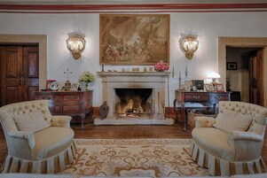 Uma sala de estar tradicional com lareira, duas poltronas, um armário de madeira e um quadro grande sobre a lareira.