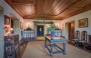 Quarto tradicional com vigas de madeira no teto, uma mesa central com um vaso e móveis clássicos.