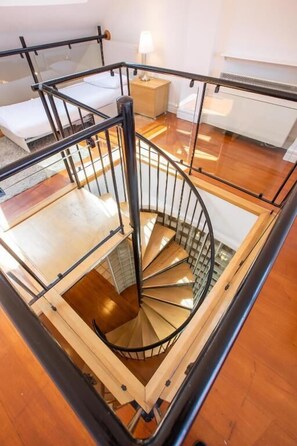 Uma escadaria em espiral com guarda-corpos de vidro e degraus de madeira, que leva a um quarto com cama e mesa de cabeceira.