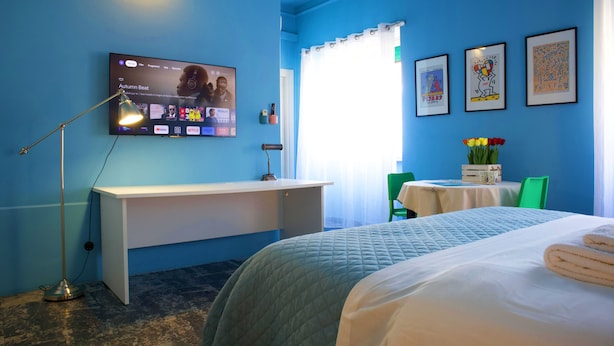 Quarto de hotel com uma cama, uma escrivaninha, uma televisão exibindo um programa e uma janela com cortinas.