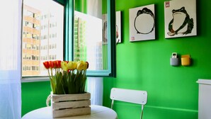 Quarto com parede verde, janela com vista para edifícios, mesa com toalha branca, caixa de madeira com flores e obras de arte na parede.