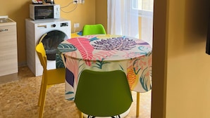 Quarto compacto com máquina de lavar, micro-ondas, uma pequena mesa com toalha colorida e uma cadeira verde.