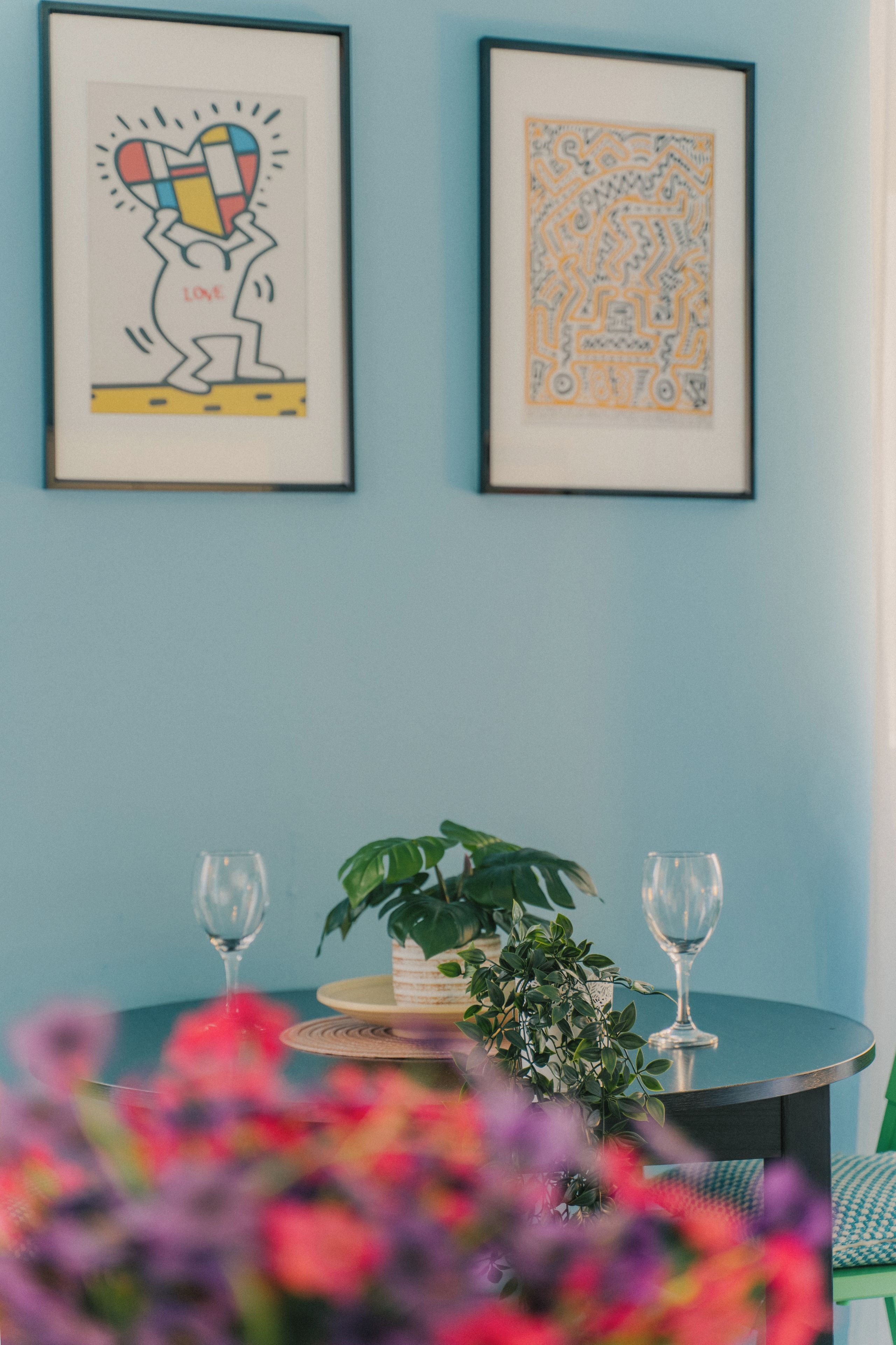 Quarto com duas obras de arte emolduradas na parede, uma mesa redonda pequena com uma planta em vaso e duas taças de vinho, e vista para uma parede azul.