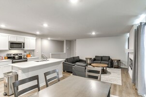 Uma cozinha moderna com armários brancos, micro-ondas e fogão. Há uma sala de jantar com mesa e cadeiras, e um sofá cinza na sala de estar.