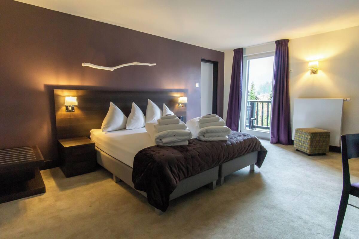 Quarto de hotel com uma cama grande, duas mesas de cabeceira com luminárias, um pequeno banquinho e vista para o exterior através de uma janela com cortinas roxas.