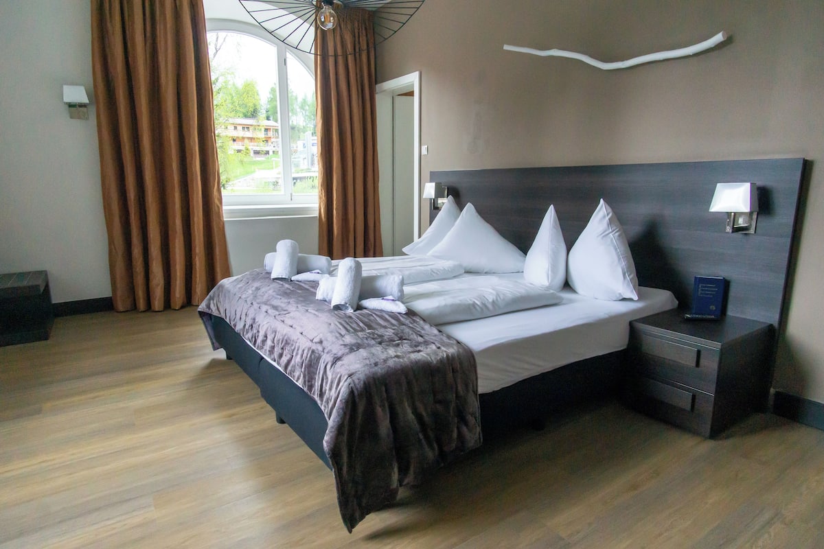 Quarto de hotel com uma cama grande, uma mesa de cabeceira, um abajur e vista para o exterior pela janela.