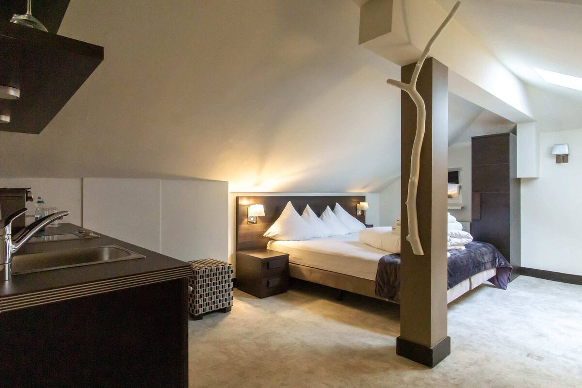 Quarto de hotel moderno com uma cama grande, escrivaninha embutida com pia e vista da cidade através de um claraboia.