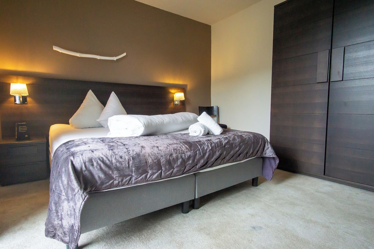 Quarto de hotel moderno com uma cama grande, duas luminárias de cabeceira, um guarda-roupa de madeira escura e esquema de cores neutras.