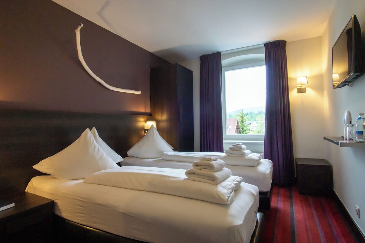 Quarto de hotel com duas camas, uma janela com cortinas, uma TV de tela plana e uma pequena mesa com uma garrafa e copos.