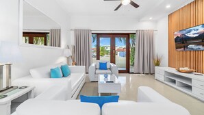 Uma sala de estar moderna com ventilador de teto, sofás brancos com almofadas azuis, uma mesa de centro e uma TV em uma parede de madeira.