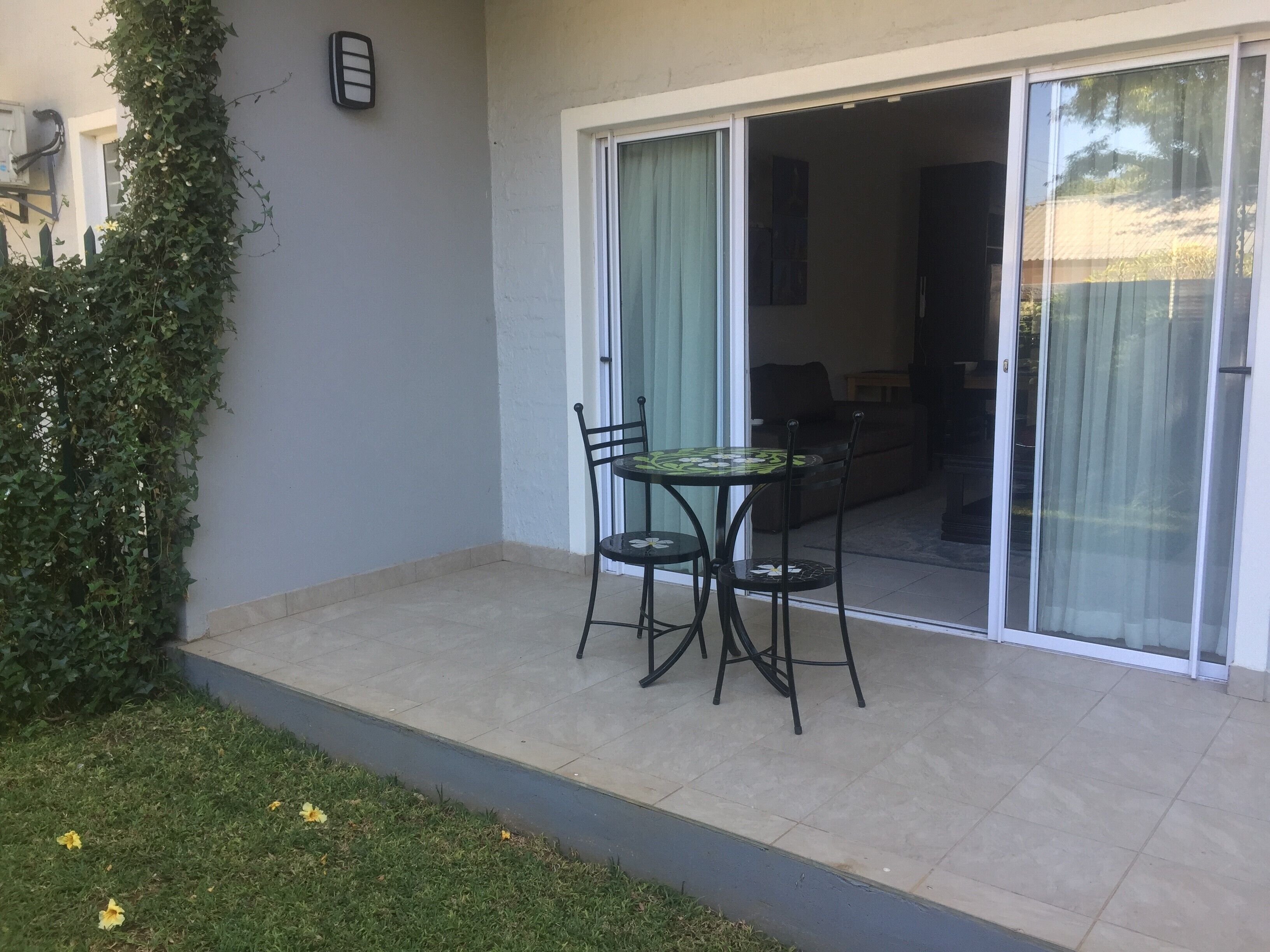 Um pátio com mesa e cadeiras, uma porta de vidro deslizante que dá para um espaço interno e uma pequena área de jardim com grama e flores.