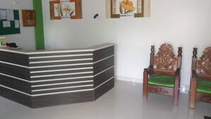 Uma área de recepção com um balcão moderno, duas cadeiras de madeira com almofadas verdes e arte floral emoldurada na parede.