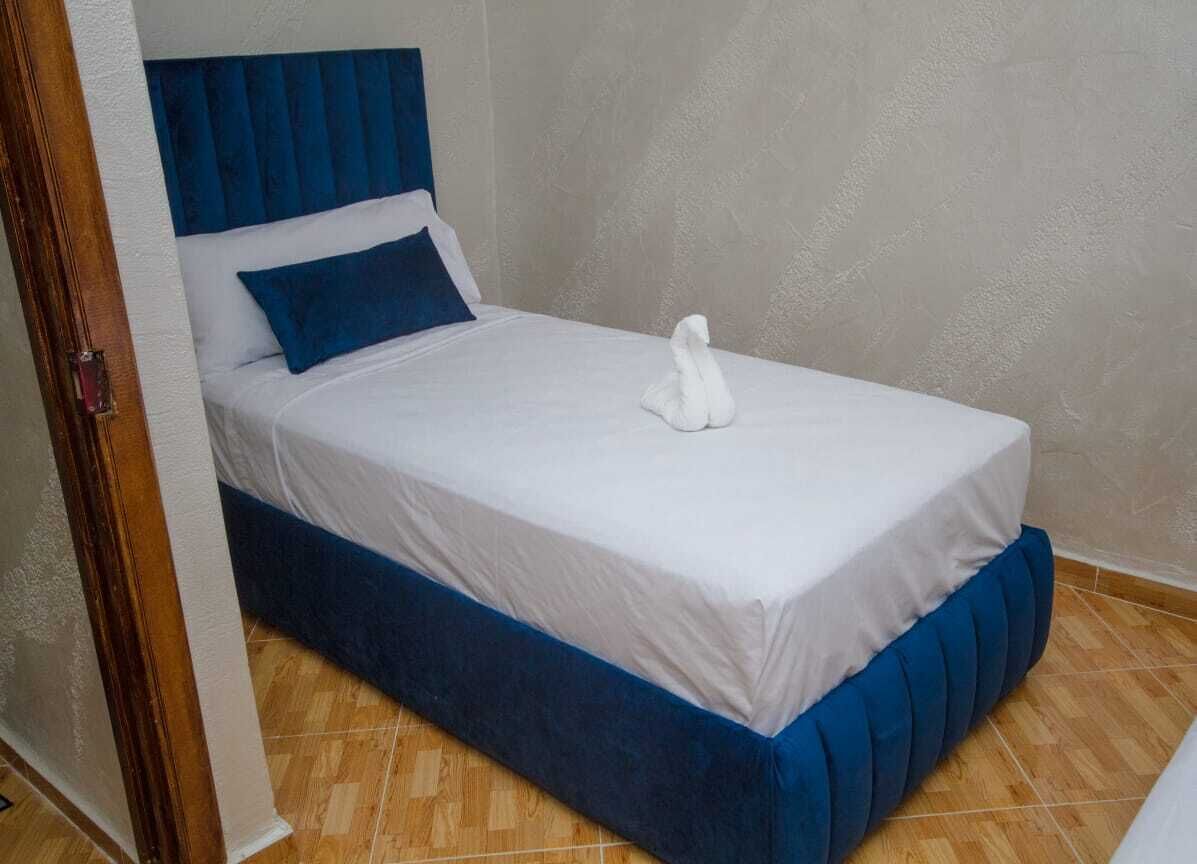 Uma cama bem arrumada com cabeceira e peseira azuis, edredom branco e uma toalha branca dobrada sobre a cama.
