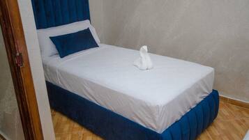 Uma cama bem arrumada com cabeceira e peseira azuis, edredom branco e uma toalha branca dobrada sobre a cama.