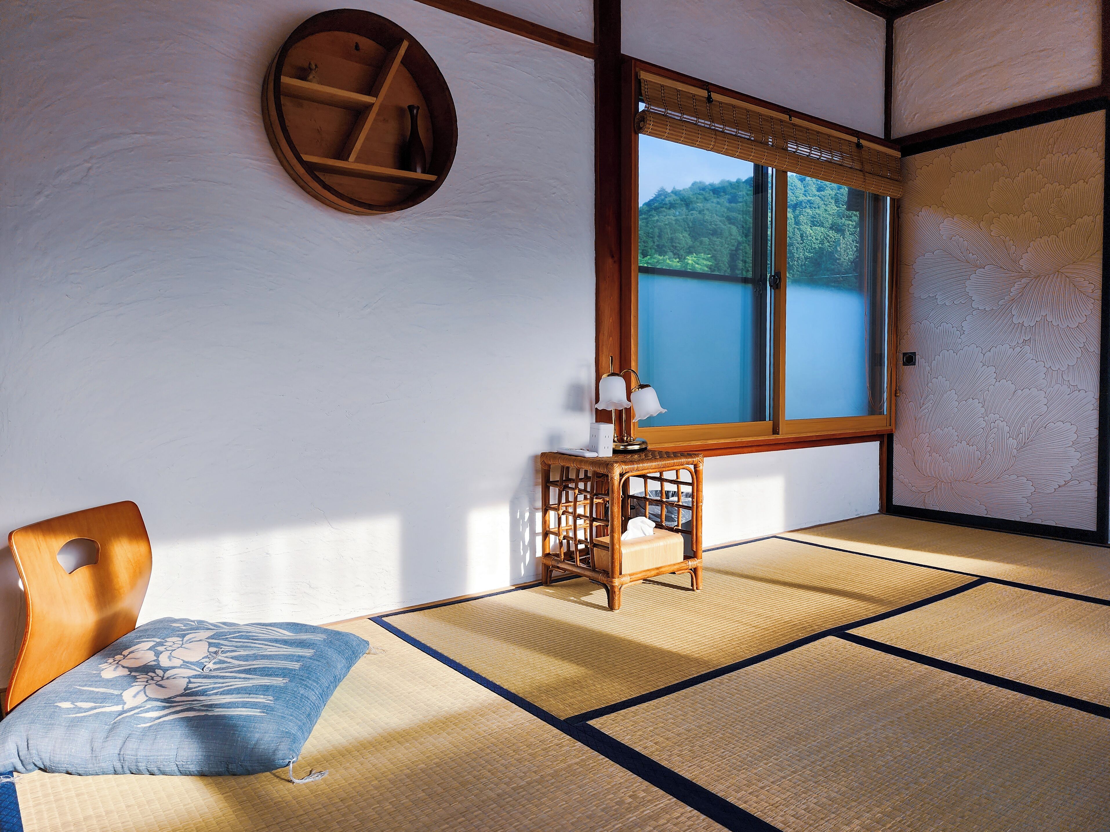 Um quarto japonês tradicional com tatames, uma mesa de madeira com uma luminária, uma cadeira e uma janela com vista para uma área arborizada.
