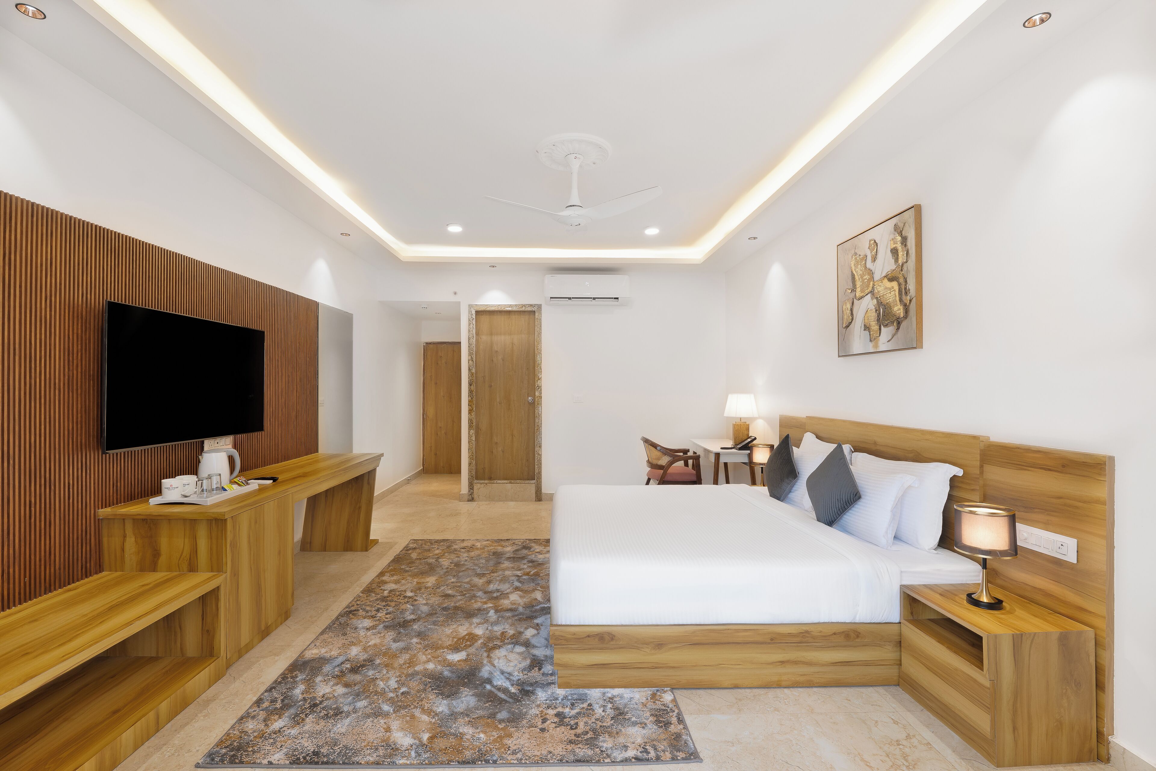 Quarto de hotel moderno com uma cama de madeira, televisão de tela plana, escrivaninha e mesa de cabeceira com luminária.