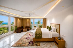 Quarto de hotel com uma cama grande, varanda com vista para o oceano, área de estar com sofá e poltronas, e uma pequena mesa com uma planta.