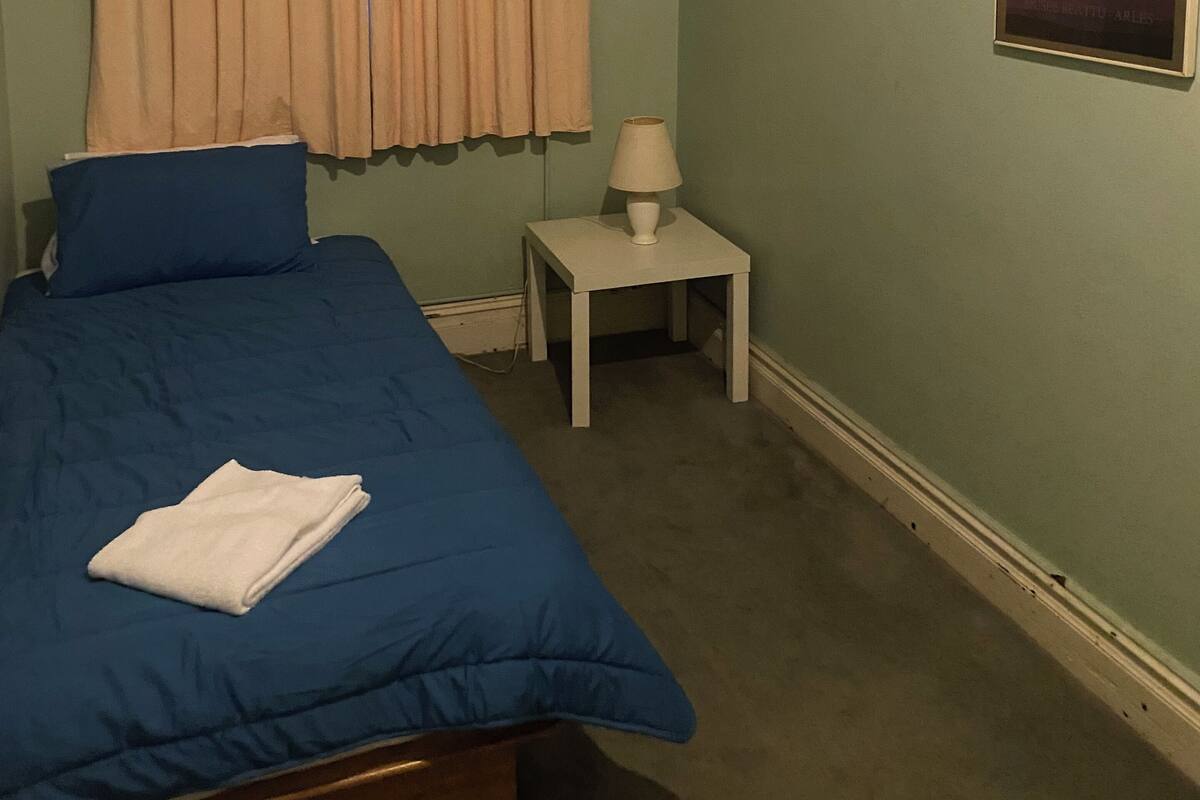 Quarto com cama, mesa de cabeceira, abajur, janela com cortinas e um quadro na parede.