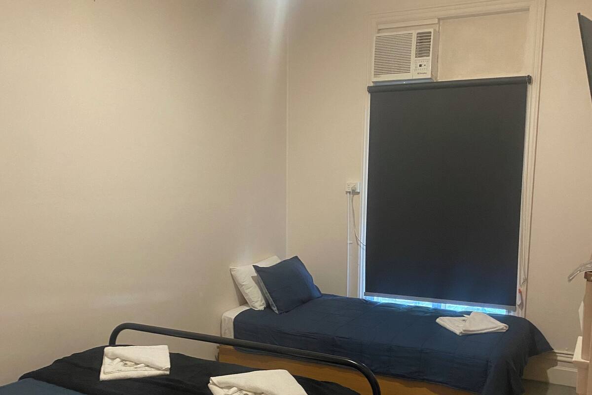 Quarto pequeno e com pouca luz, com duas camas de solteiro, cada uma com colchão preto e almofadas brancas. Há uma janela com persianas fechadas e uma luminária no teto visível.