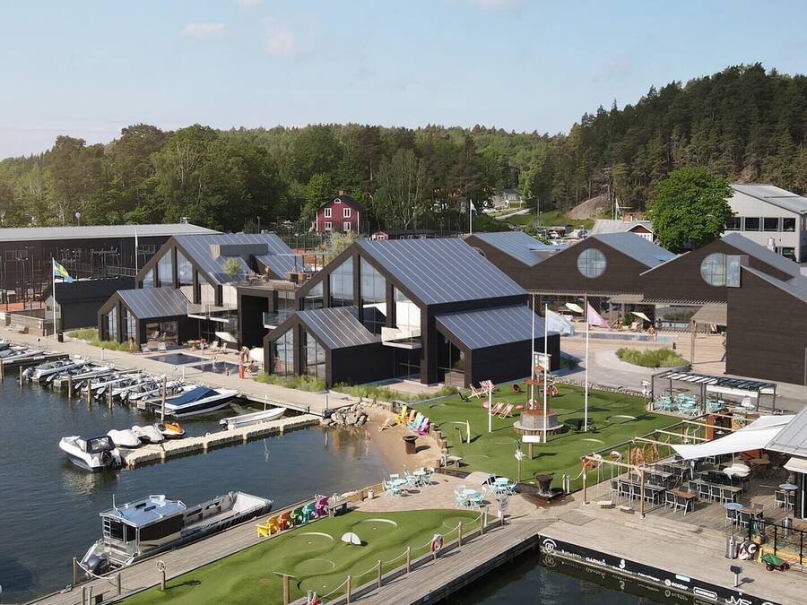 Uma marina com barcos atracados, edifícios modernos com grandes janelas e uma área gramada com mesas ao ar livre.