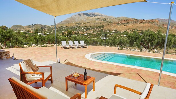 Uma área à beira da piscina com espreguiçadeiras, uma mesa com garrafa e copos, e vista para montanhas.