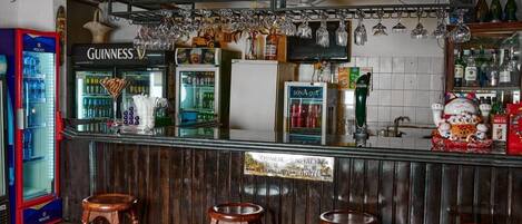 Uma área de bar com banquetas, um refrigerador Guinness e várias garrafas e copos em prateleiras.