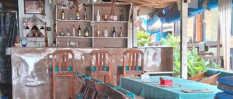 Uma área de bar com cadeiras e mesas de madeira, um balcão com garrafas e luminárias pendentes.
