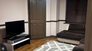 Uma sala de estar com uma TV de tela plana em um móvel, um sofá marrom escuro, um tapete estampado e uma porta com um vitral acima.