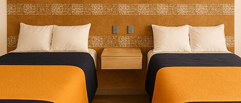 Quarto de hotel com duas camas, cada uma com colcha azul marinho e laranja, cabeceira de madeira com padrão decorativo e pequena mesa de cabeceira com dois interruptores de luz.