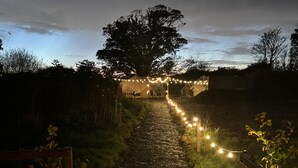 Uma trilha conduz a uma área com tendas, iluminada por luzes penduradas, cercada por árvores e vegetação.