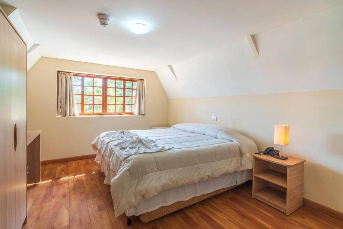Quarto com cama, mesa de cabeceira com abajur, janela com cortinas e piso de madeira.