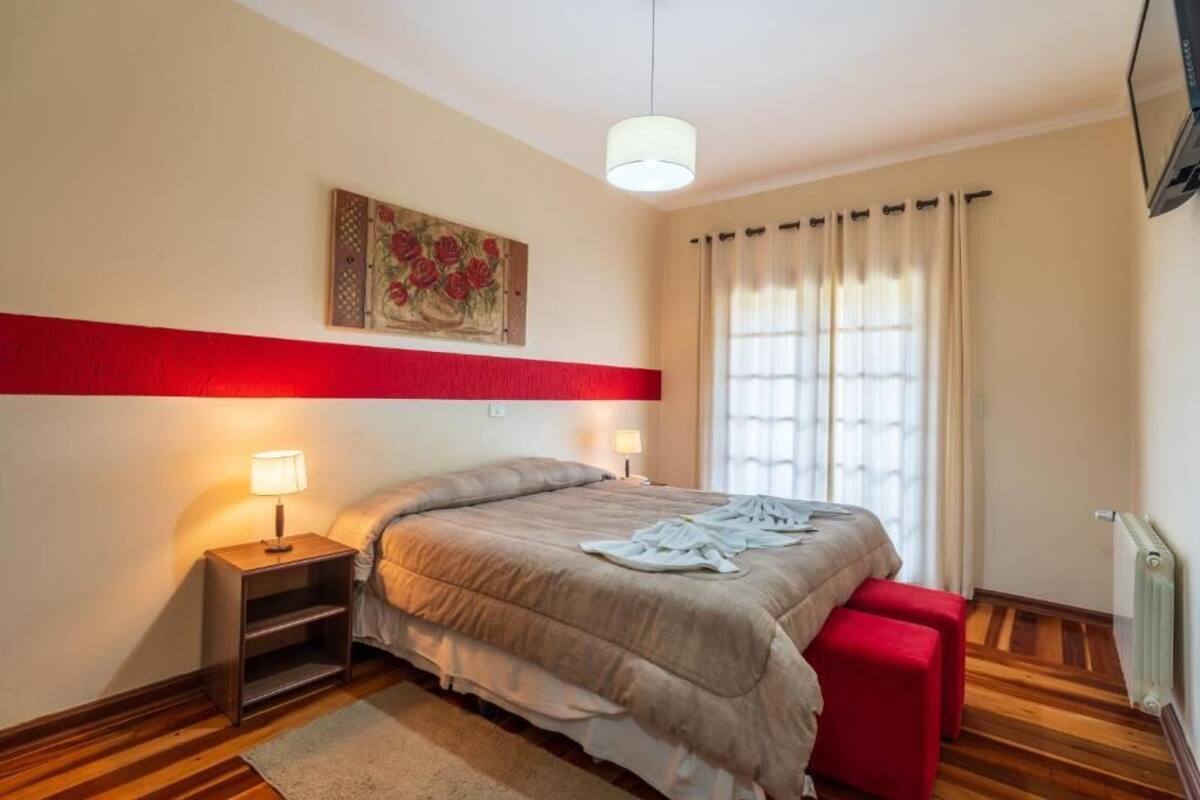 Quarto com cama, abajures, parede em destaque vermelha, janela com cortinas e radiador.